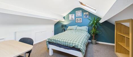 Quarto com teto inclinado, cama com colcha estampada, escrivaninha, cadeira, radiador, planta em vaso e prateleira de madeira.