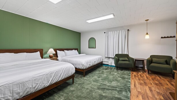 Quarto de hotel com duas camas, uma mesa pequena, duas poltronas, um tapete verde e piso de madeira.