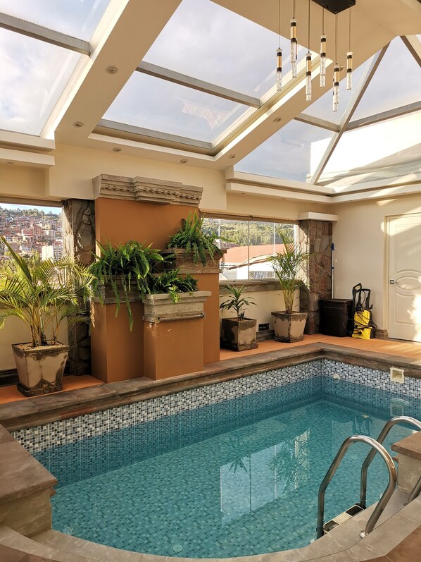 Piscina com teto de vidro, cercada por plantas em vasos, e vista para a paisagem urbana.