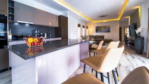 Uma cozinha moderna com uma ilha central, uma área de refeições com banquetas, uma sala de estar com sofá e TV, e uma janela grande.