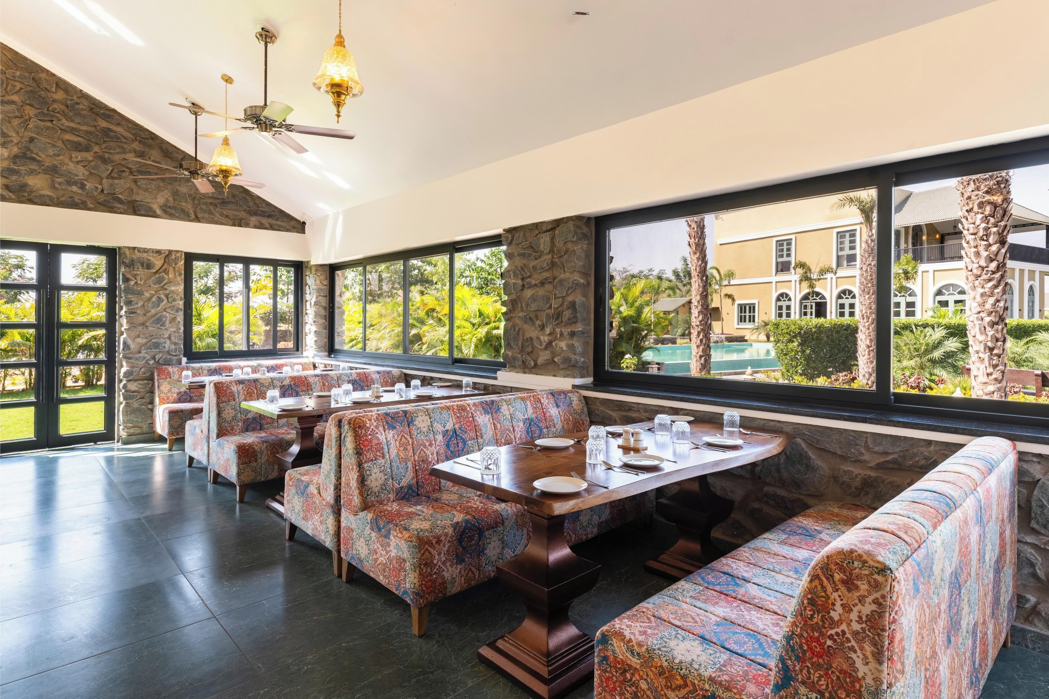 Restaurante com tetos altos, paredes de pedra e amplas janelas com vista para uma piscina e vegetação.