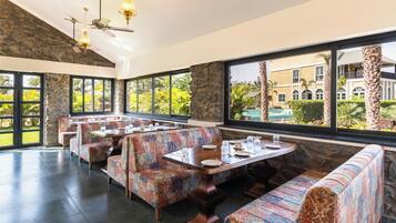 Restaurante com tetos altos, paredes de pedra e amplas janelas com vista para uma piscina e vegetação.