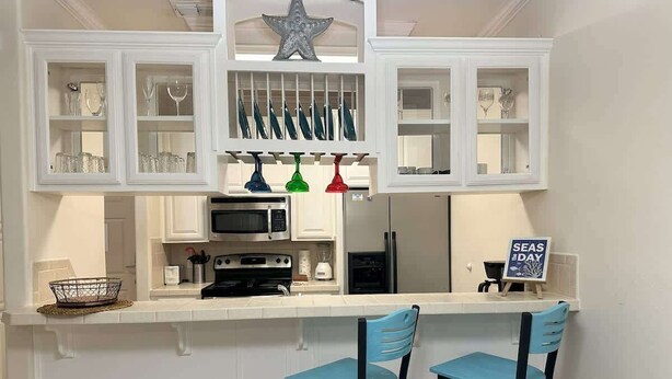 Cozinha com armários brancos, micro-ondas, geladeira e dois banquetas azuis.