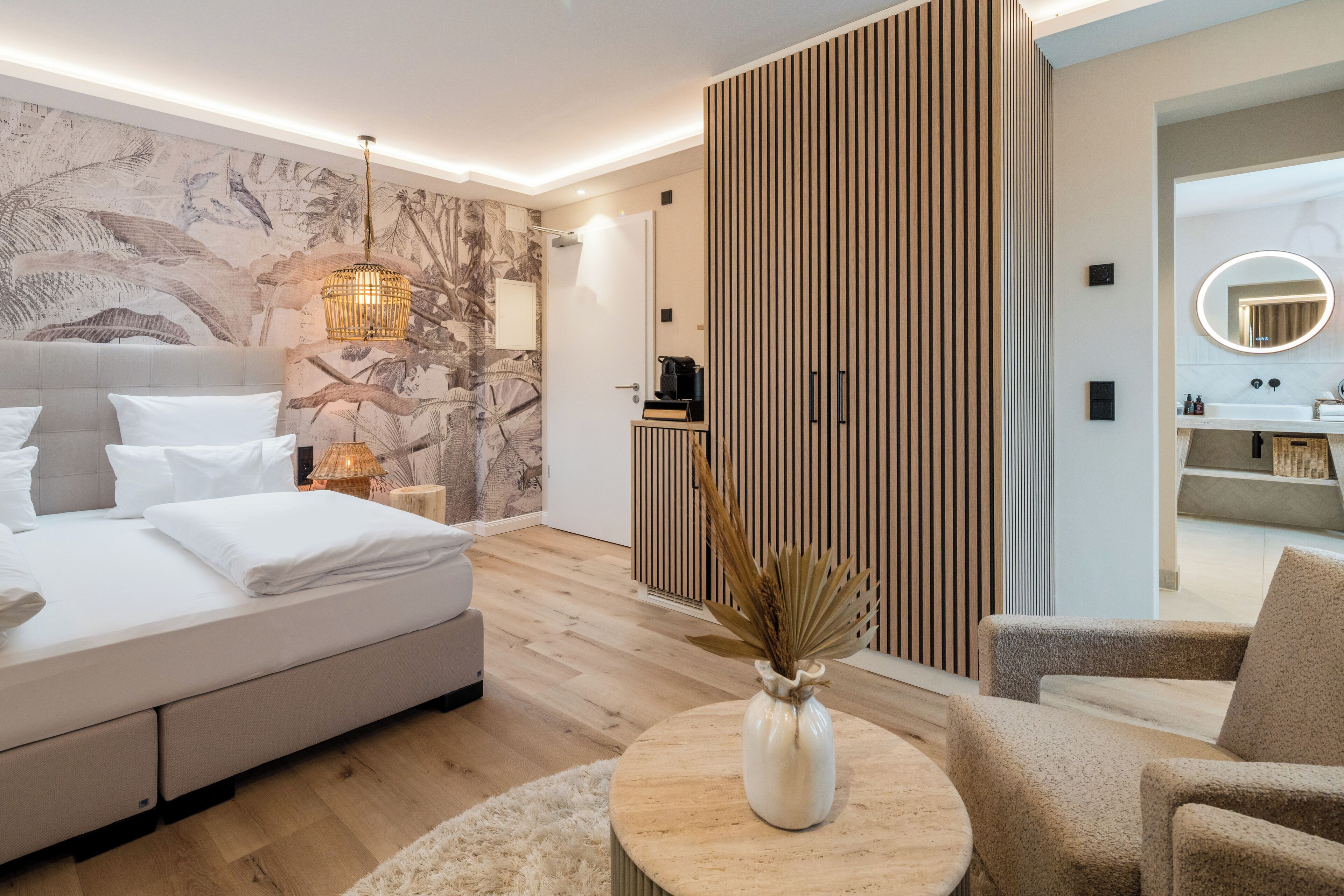 Quarto de hotel moderno com cama, mesa de centro redonda, área de estar, parede com mural tropical e divisória de madeira.
