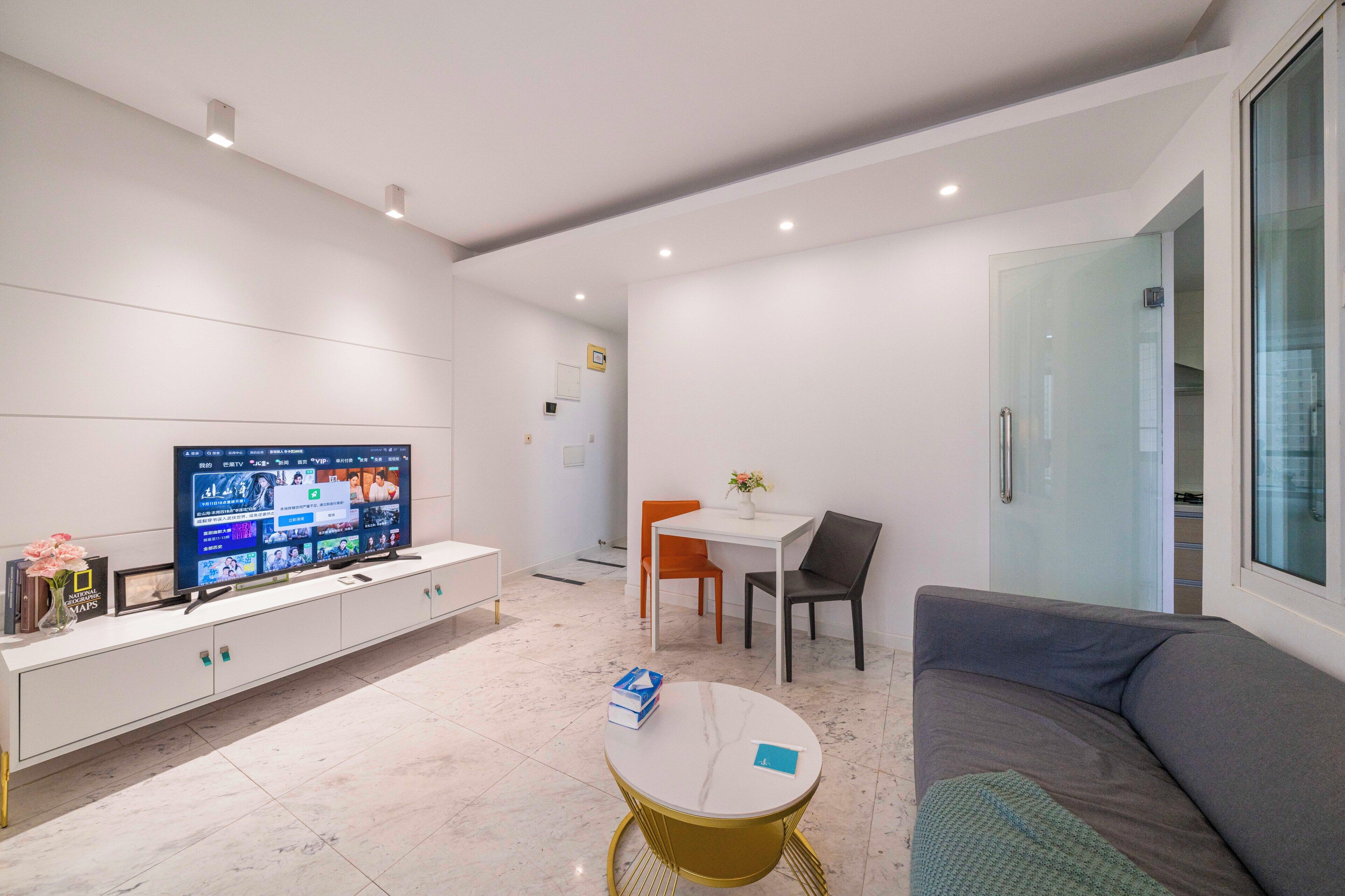 Uma sala de estar moderna com uma televisão de tela plana, um sofá, uma mesinha redonda e uma área de jantar com uma mesa e cadeiras brancas.