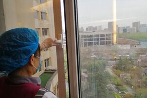 Uma pessoa com cobertura azul na cabeça e colete vermelho parada perto de uma janela, olhando para o exterior.