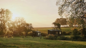 Paisagem rural com duas pequenas cabanas rodeadas por árvores e campos abertos.