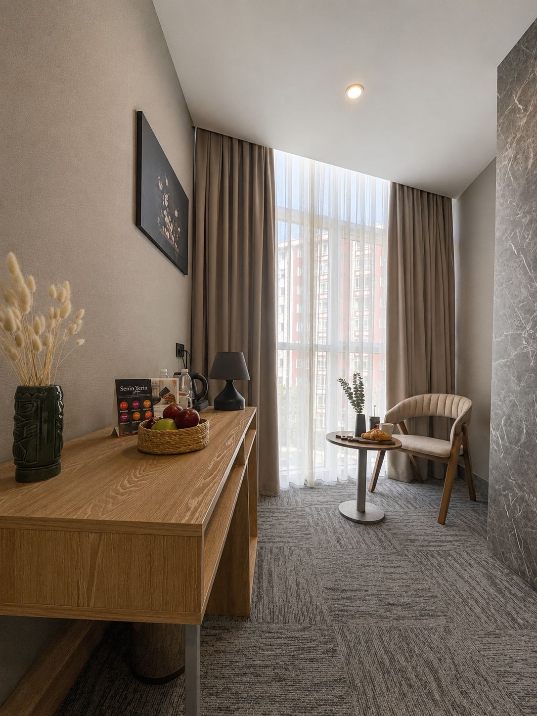 Quarto de hotel com escrivaninha de madeira, uma cadeira, uma pequena mesa com um prato de comida, um vaso com flores secas e uma moldura com um quadro na parede.