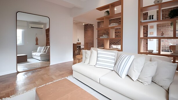 Uma sala de estar moderna com um sofá branco, um espelho grande e estantes de madeira embutidas com diversos objetos.