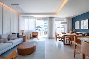 Quarto de hotel moderno com sofá, mesa de jantar, cadeiras e uma grande janela com vista para a cidade.