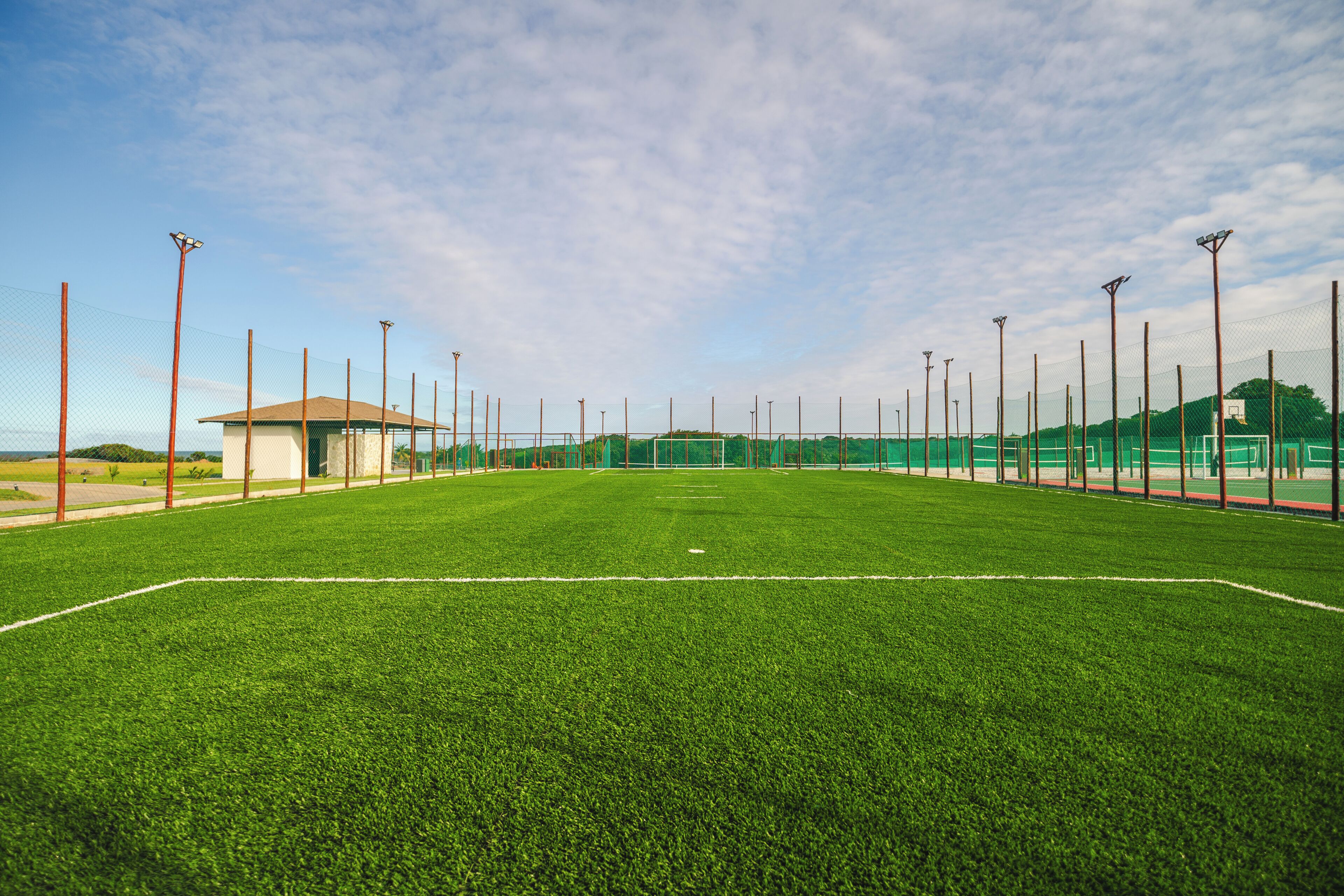 Um campo de esportes ao ar livre com postes de gol, refletores e um pequeno edifício, provavelmente um vestiário ou depósito.