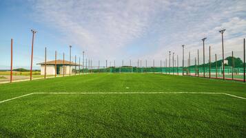 Um campo de esportes ao ar livre com postes de gol, refletores e um pequeno edifício, provavelmente um vestiário ou depósito.