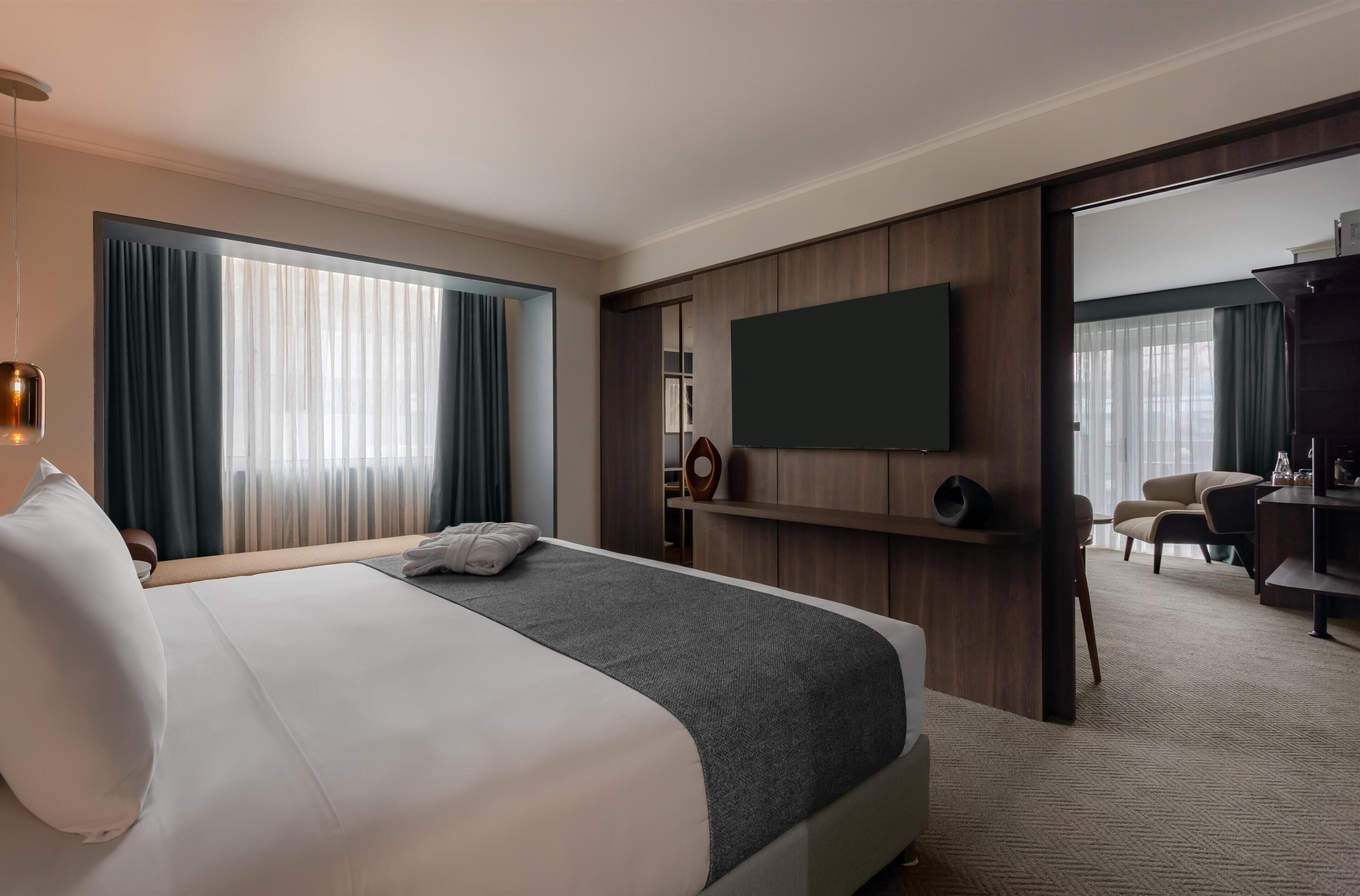 Quarto de hotel com uma cama grande, televisão de tela plana na parede, escrivaninha com cadeira e vista para a cidade pela janela.