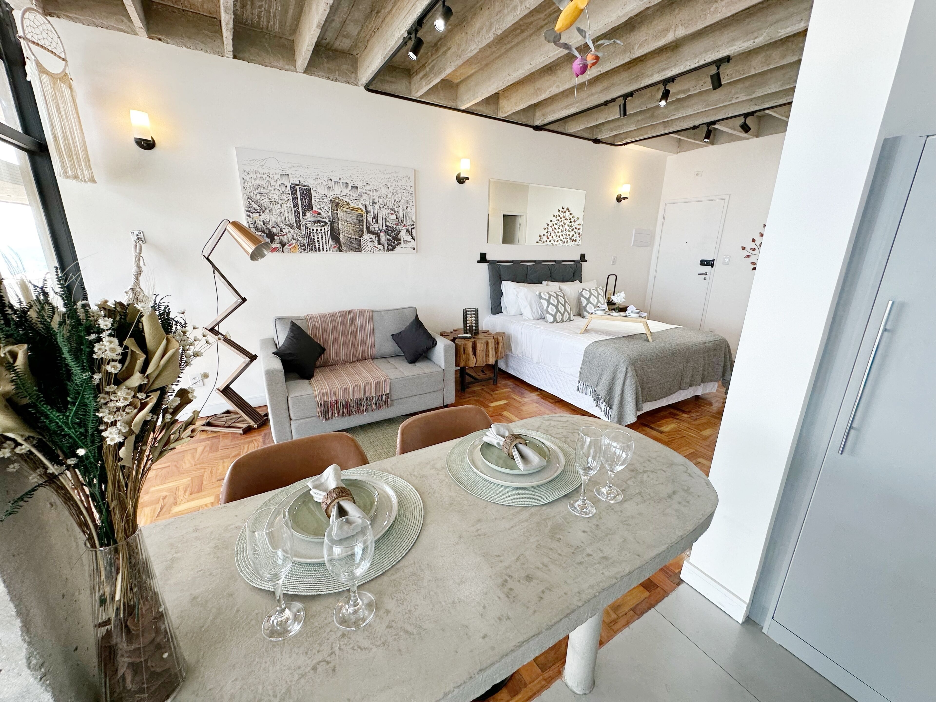 Decoração moderna com mesa de jantar posta para dois, um sofá, uma cama e uma pintura com vista para a cidade.