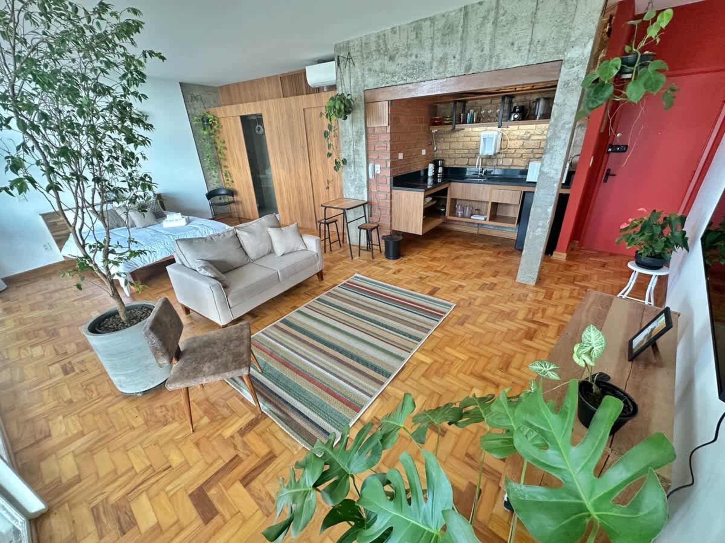 Um espaço moderno com sofá, mesa de centro e área de jantar. Há uma cozinha com uma parede de destaque vermelha e um vaso de planta.