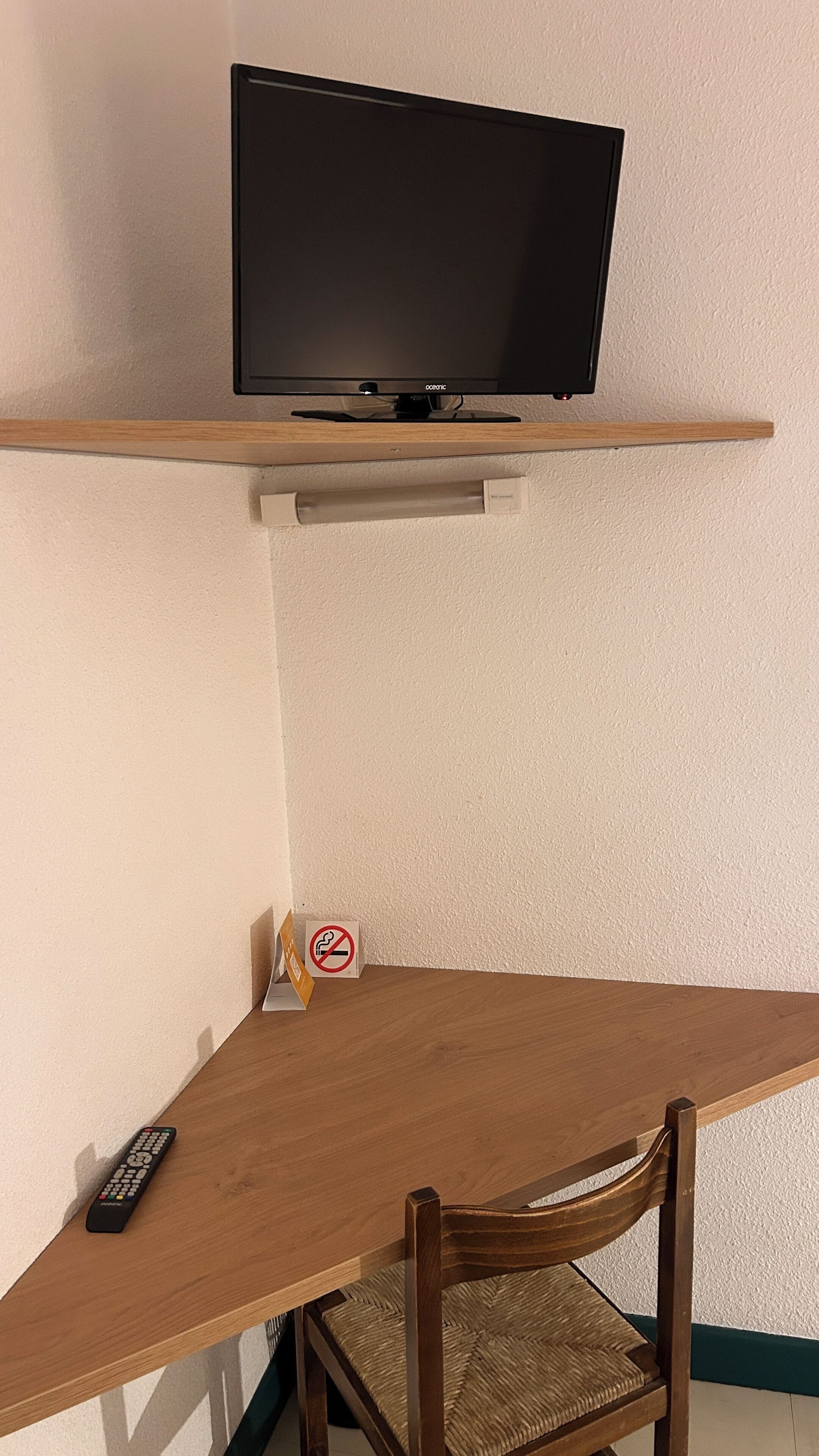 Quarto de canto com televisão fixada numa prateleira de madeira, uma secretária com uma cadeira e um aviso de proibido fumar.