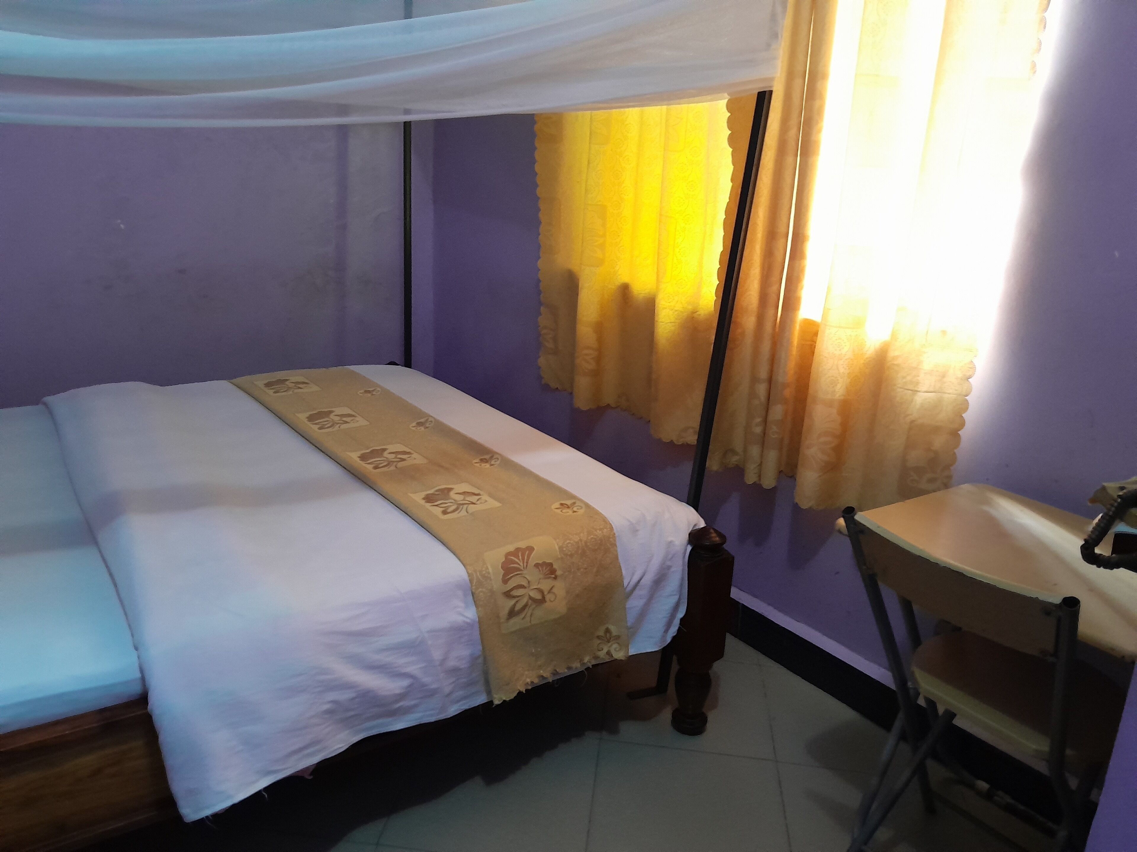 Uma cama de solteiro com colcha branca e bege, uma pequena mesa de escritório com cadeira e uma janela com cortinas amarelas.