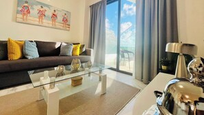 Uma sala de estar moderna com uma mesa de centro de vidro, um sofá com almofadas coloridas, um quadro com cenas de praia e vista através de grandes janelas.
