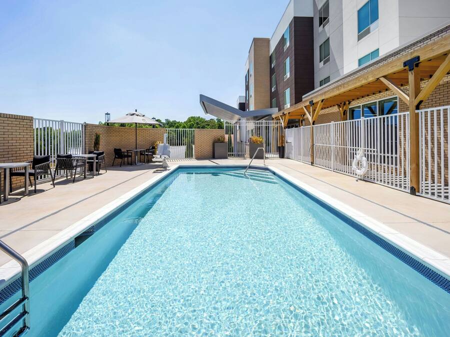 Uma piscina retangular com borda azulejada, cercada por uma cerca branca, e adjacente a um edifício com fachada de tijolos e janelas.