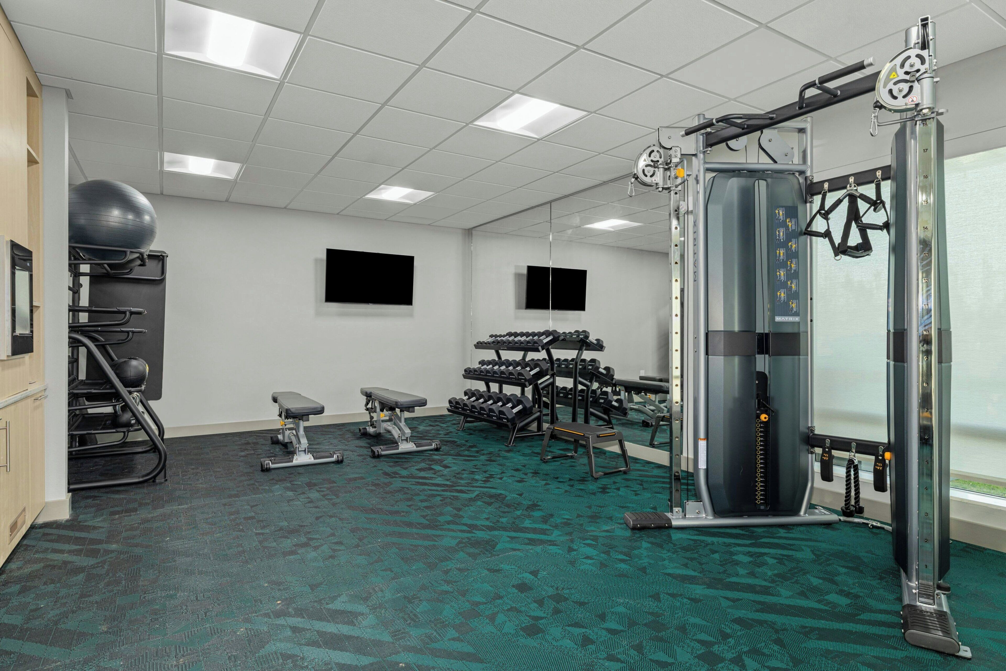 Uma academia com equipamentos de exercício, um banco, halteres e uma televisão presa na parede.