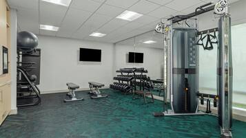 Uma academia com equipamentos de exercício, um banco, halteres e uma televisão presa na parede.