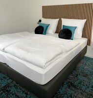 Cama com roupa de cama branca, dois travesseiros verde-água e um travesseiro de destaque preto. A cabeceira apresenta um design de painel de madeira vertical.