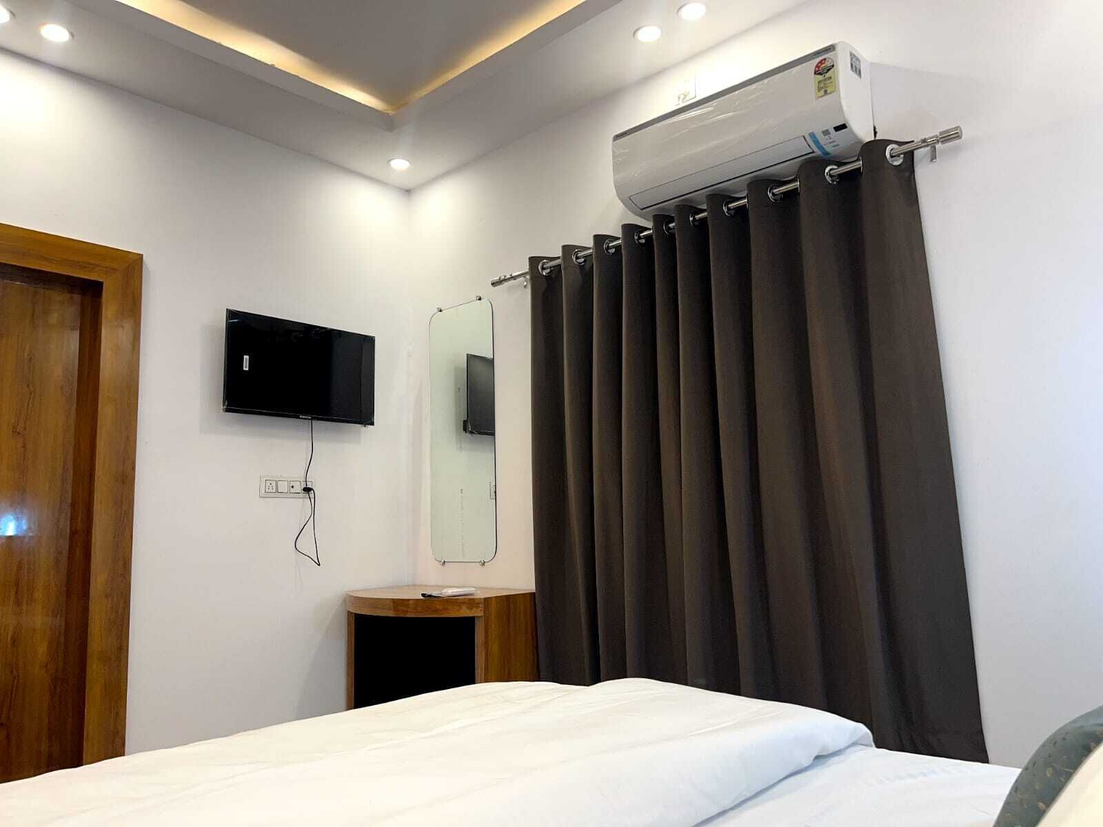 Quarto de hotel com cama, televisão na parede, espelho, janela com cortinas e unidade de ar condicionado.