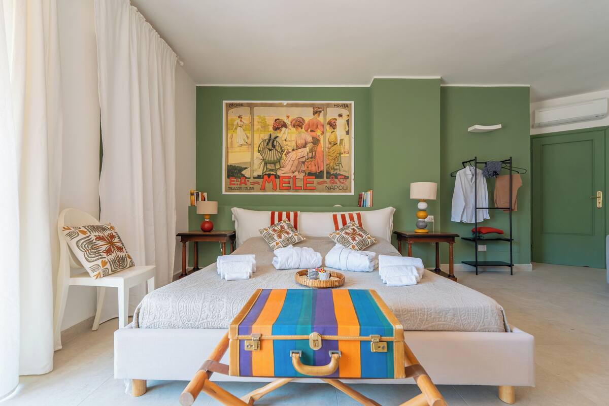 Um quarto com uma cama grande, uma mala colorida, um pôster na parede e um cabideiro.
