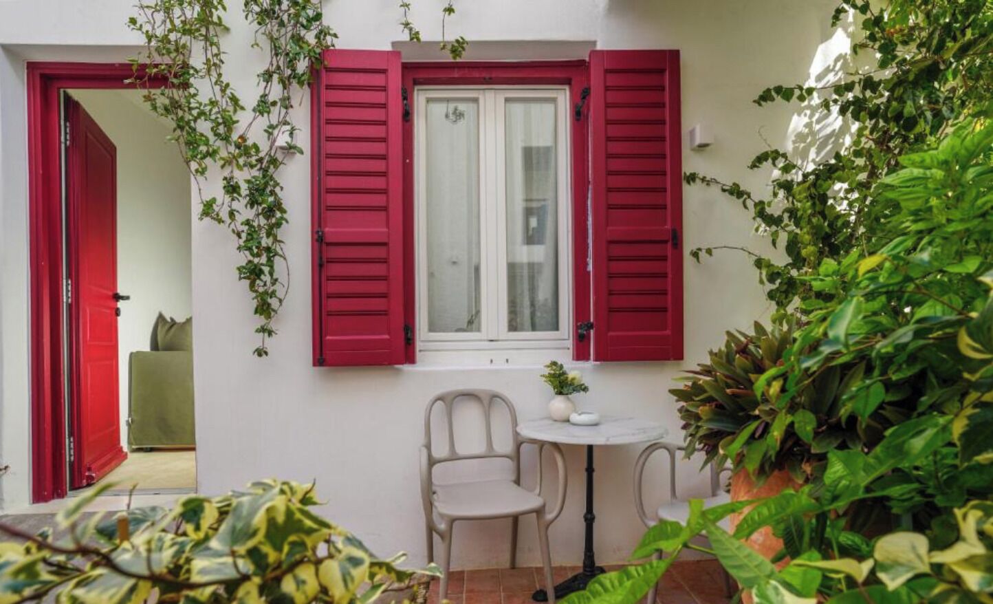 Um pequeno pátio com mesa e cadeiras, uma porta vermelha e uma janela com persianas vermelhas.