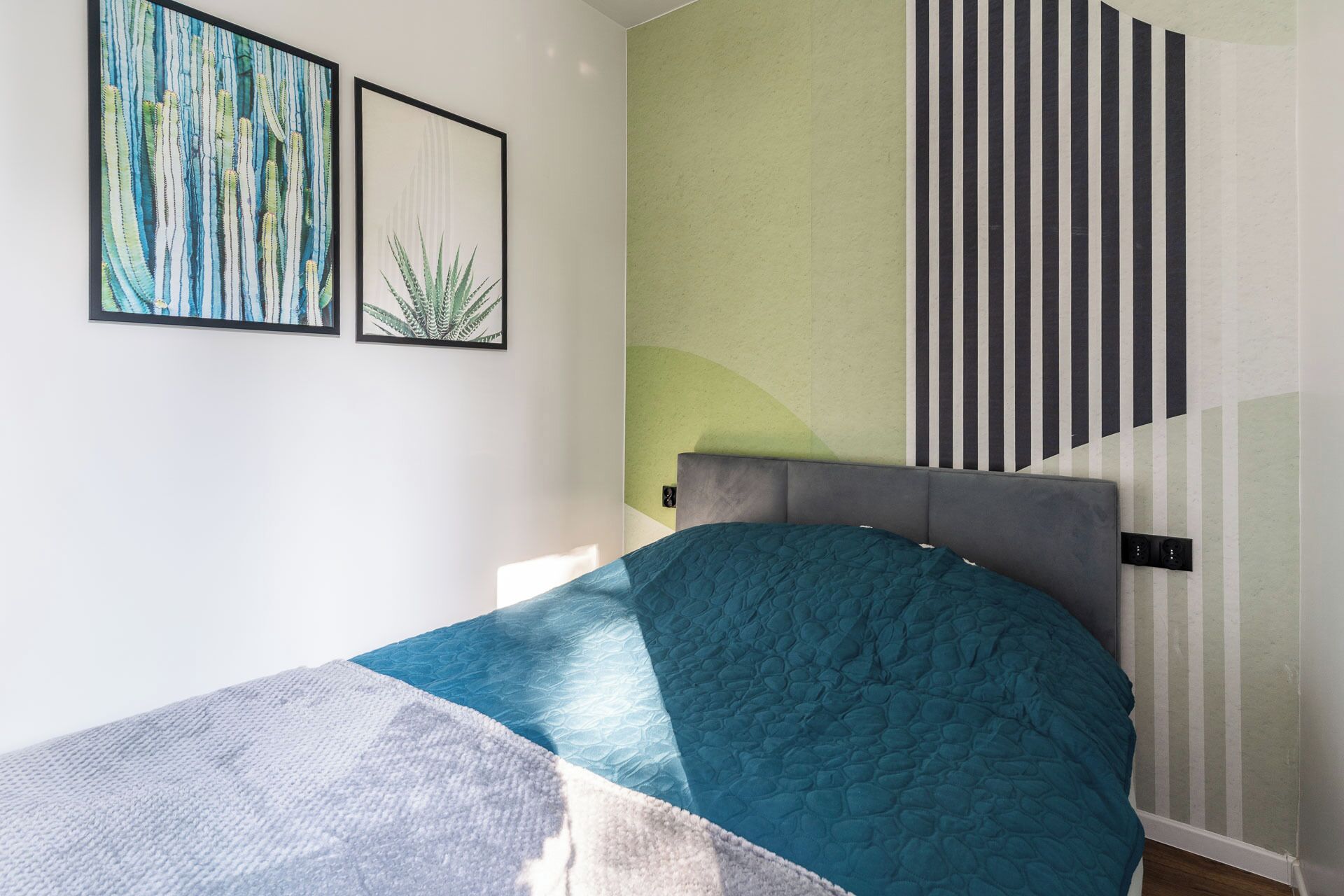 Quarto moderno com cama, TV fixada na parede e dois quadros com gravuras de plantas.