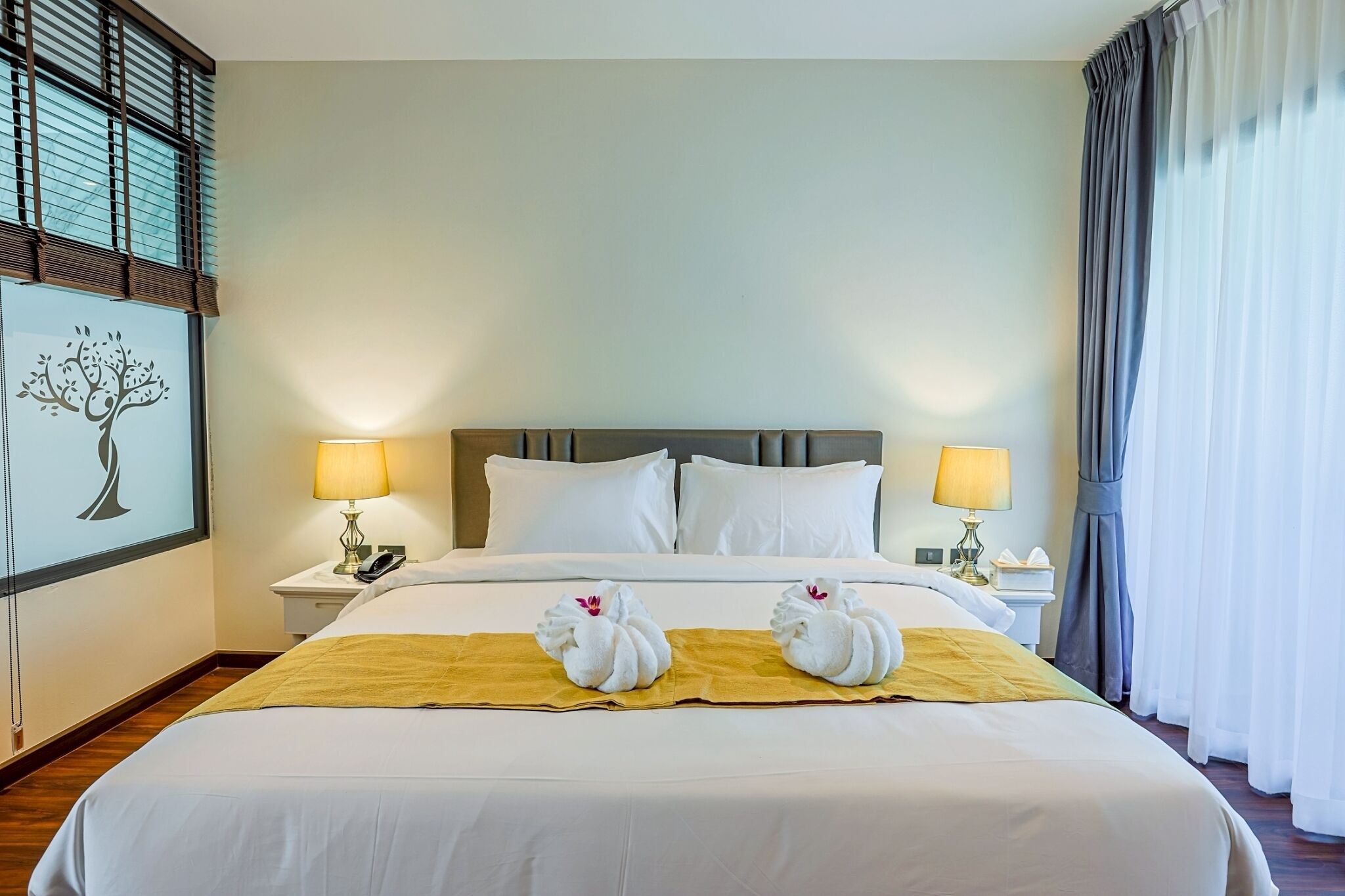 Quarto de hotel com uma cama grande, duas luminárias de cabeceira, uma mesa de cabeceira com um telefone e uma parede com uma obra de arte de uma árvore.