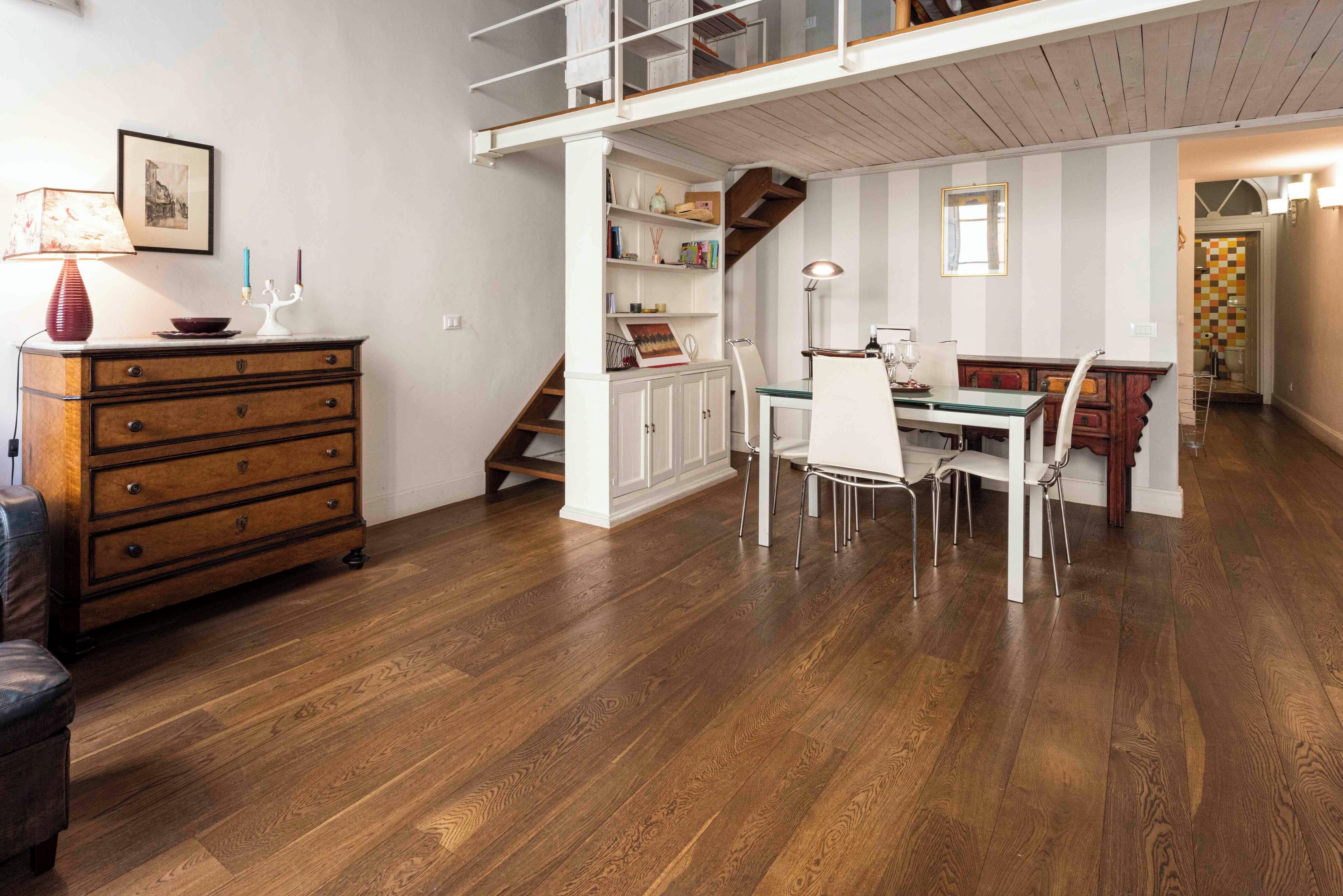 Uma sala de estar espaçosa com piso de madeira, uma mesa de jantar com cadeiras, um armário branco e uma escada que dá para um andar superior.