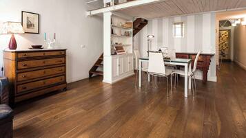 Uma sala de estar espaçosa com piso de madeira, uma mesa de jantar com cadeiras, um armário branco e uma escada que dá para um andar superior.