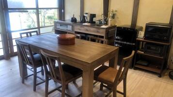 Uma mesa de jantar de madeira com cadeiras, uma máquina de café e uma televisão num quarto com janelas amplas.