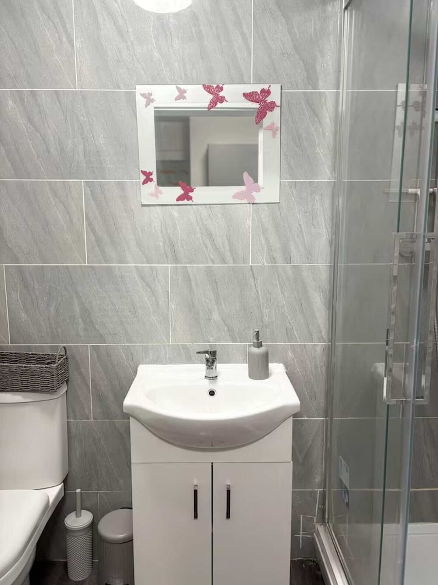 Casa de banho com lavatório branco, espelho com autocolantes de borboletas, cabine de duche e sanita.