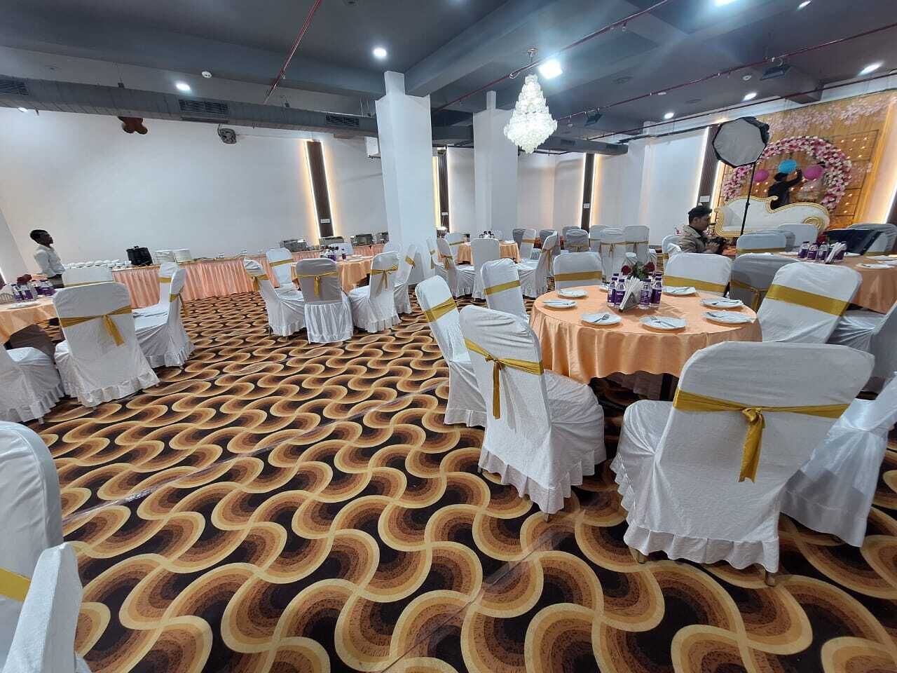 Uma sala de banquetes com mesas redondas, cadeiras brancas com fitas douradas e um tapete estampado.