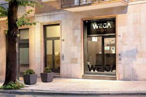 Entrada de um hotel com uma grande porta de vidro, uma placa indicando “WEGA HOTEL” e duas plantas em vasos na calçada.