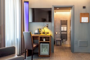 Quarto de hotel com escrivaninha, cadeira, mini-geladeira e televisão fixada na parede.
