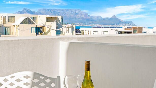 Uma varanda com uma mesa, uma garrafa de vinho e duas taças. Ao fundo, edifícios e uma montanha.
