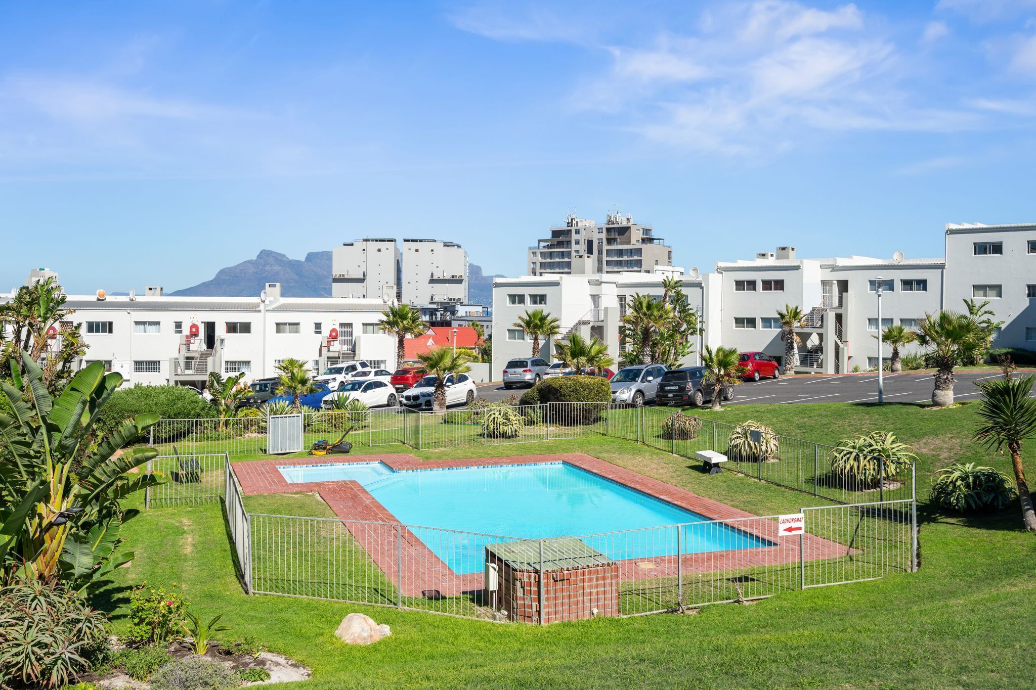 Piscina cercada por uma área vedada, com vista para edifícios modernos e carros estacionados.
