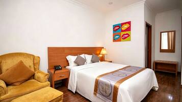 Quarto de hotel com cama, mesa de cabeceira com telefone, cadeira, mesinha de cabeceira com luminária e parede com obra de arte colorida.