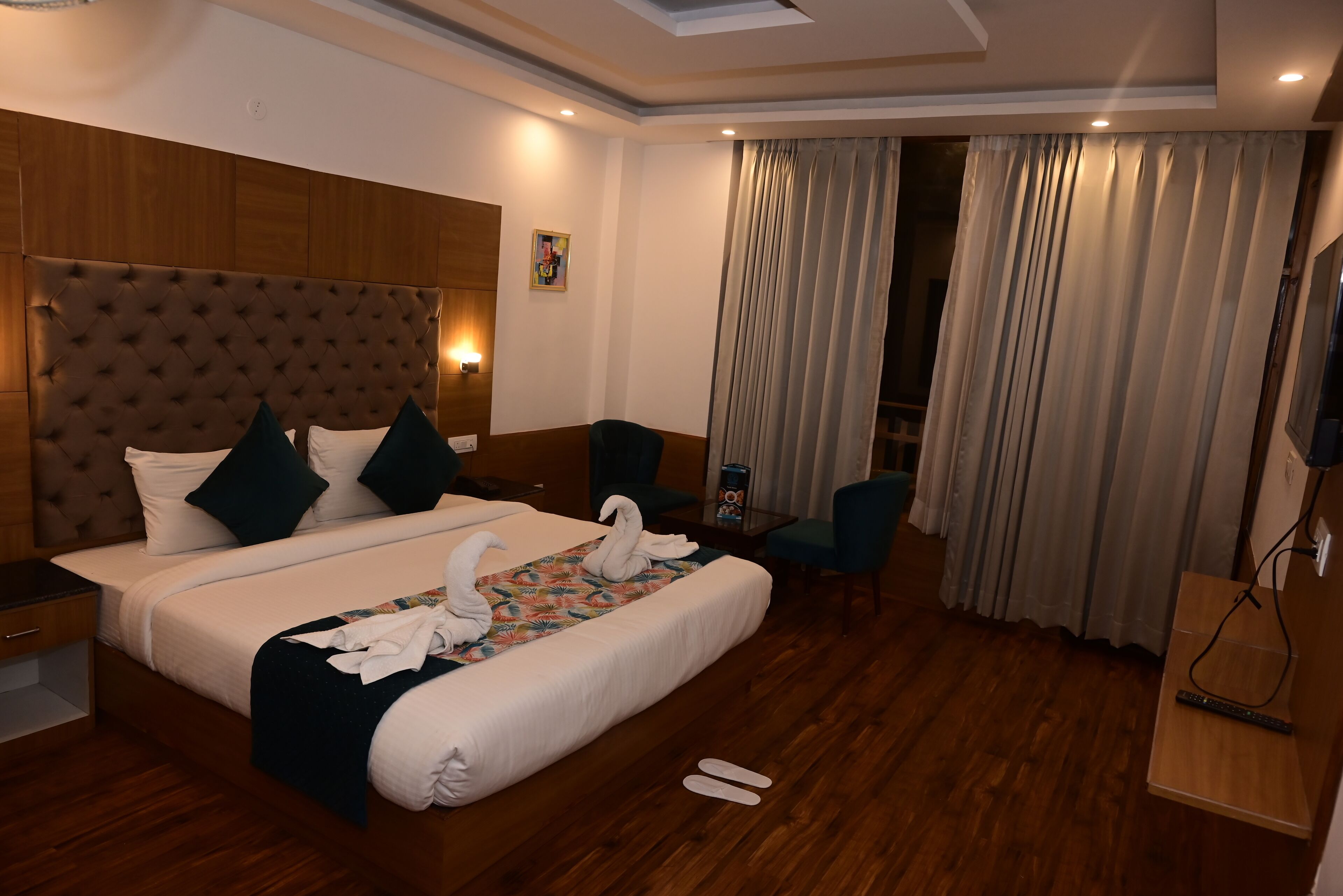 Quarto de hotel com uma cama grande, cabeceira de madeira, escrivaninha com cadeira, televisão e uma janela ampla com cortinas.