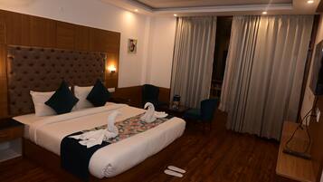 Quarto de hotel com uma cama grande, cabeceira de madeira, escrivaninha com cadeira, televisão e uma janela ampla com cortinas.