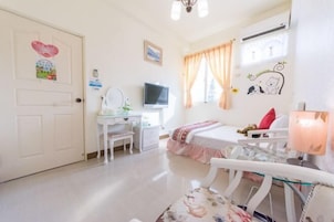 Quarto bem iluminado com cama, escrivaninha, cadeira, televisão e janela com cortinas.