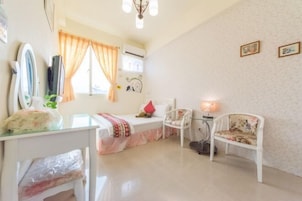 Quarto pequeno e bem iluminado, com cama, cadeira, mesa e abajur.
