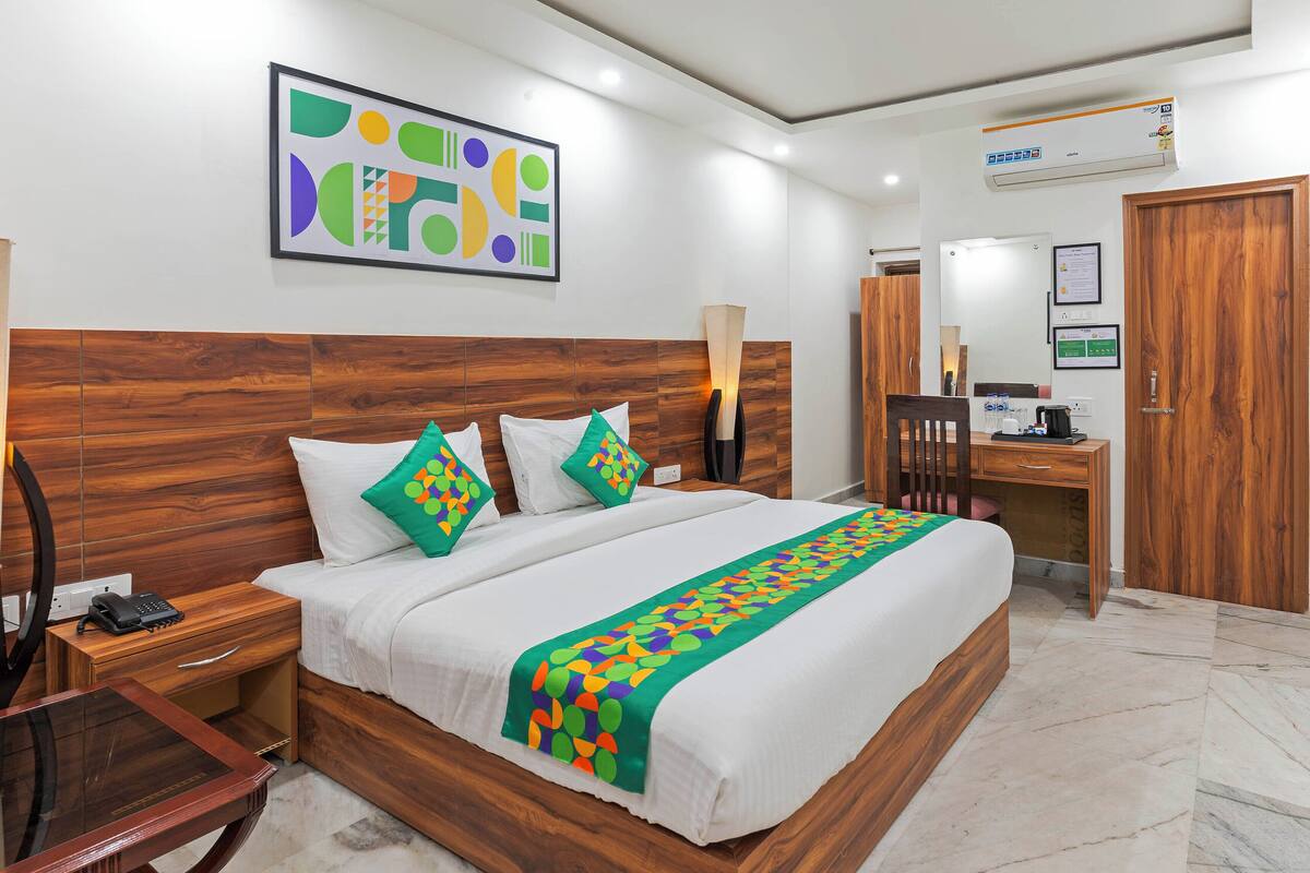 Quarto de hotel com cabeceira de madeira, cama com roupa de cama branca, manta verde e laranja, escrivaninha com cadeira e ar condicionado fixado na parede.