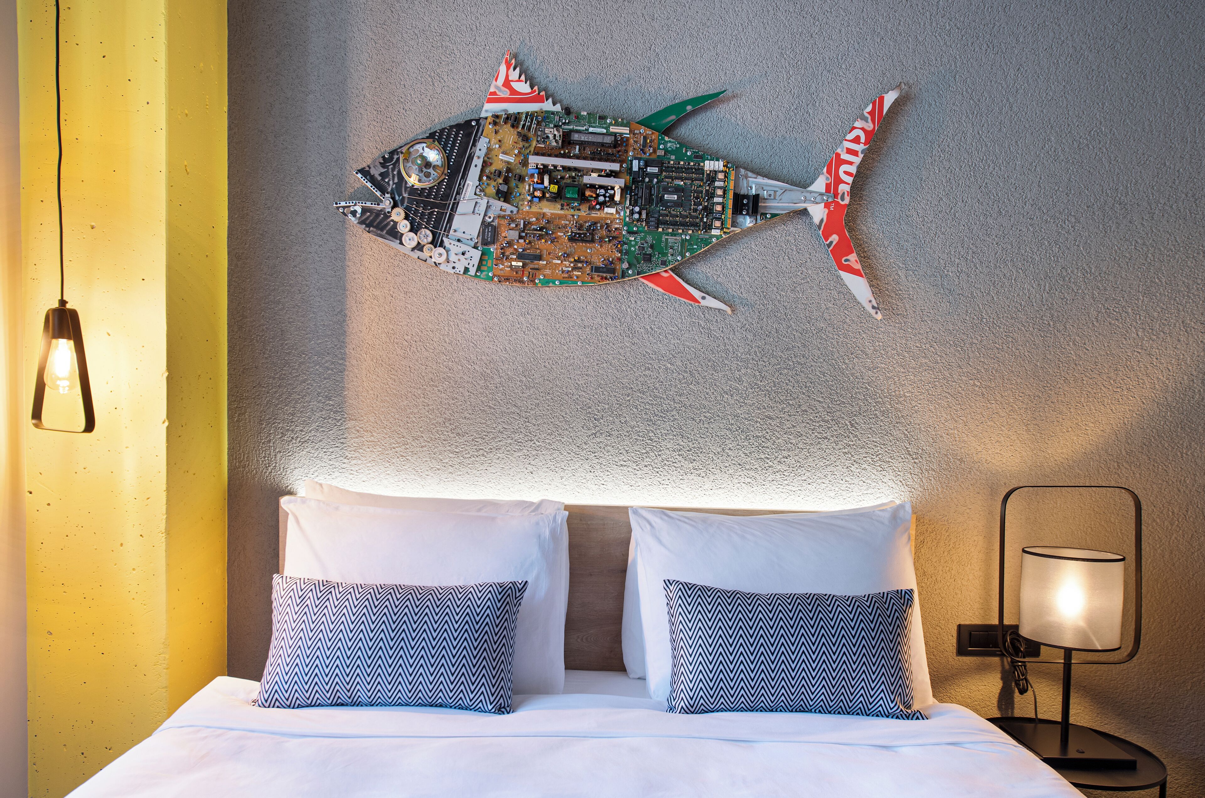 Uma cama com duas almofadas estampadas, uma luminária de cabeceira e uma obra de arte em formato de peixe na parede.