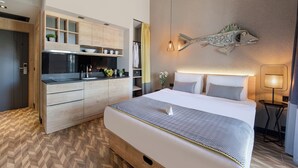 Quarto de hotel moderno com uma cama grande, cozinha planejada com armários de madeira, arte de parede com tema de peixes e uma mesinha de cabeceira.