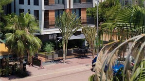 Um edifício moderno de vários andares com varandas, painéis solares e um espaço pavimentado com palmeiras.
