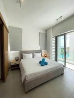 Quarto de hotel moderno com uma cama grande, uma mesa de cabeceira, um abajur e vista para uma piscina através de uma porta de vidro deslizante.